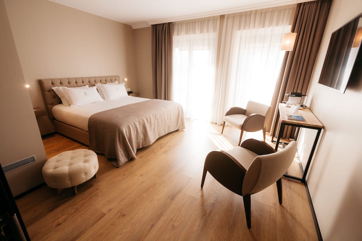 Quarto de hotel com uma cama grande, uma escrivaninha com cadeira, um banquinho pequeno e uma televisão fixada na parede.