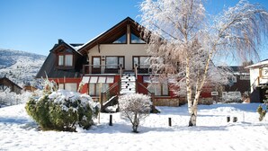 Uma casa de madeira de dois andares com telhado vermelho, rodeada por terreno e árvores cobertos de neve.