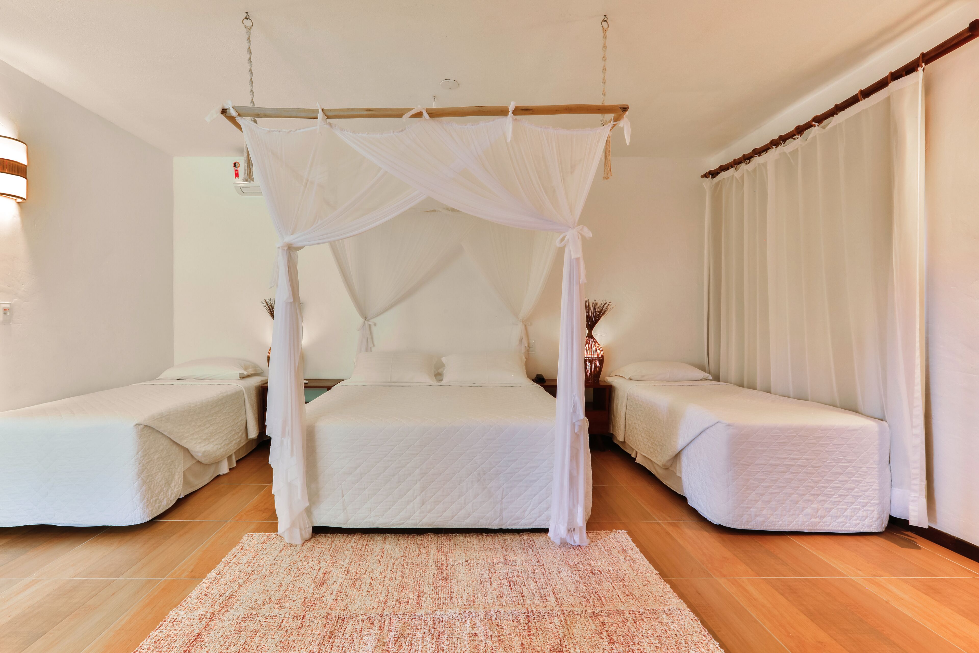 Quarto com cama de dossel, duas camas de solteiro, piso de madeira e parede branca.