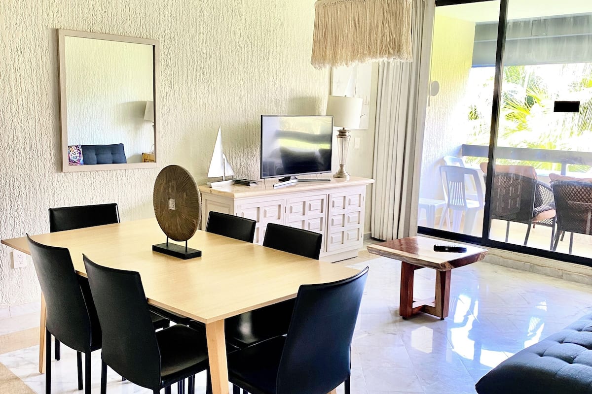 Uma sala de estar moderna com uma mesa de jantar grande, cadeiras pretas, uma televisão e um lustre.
