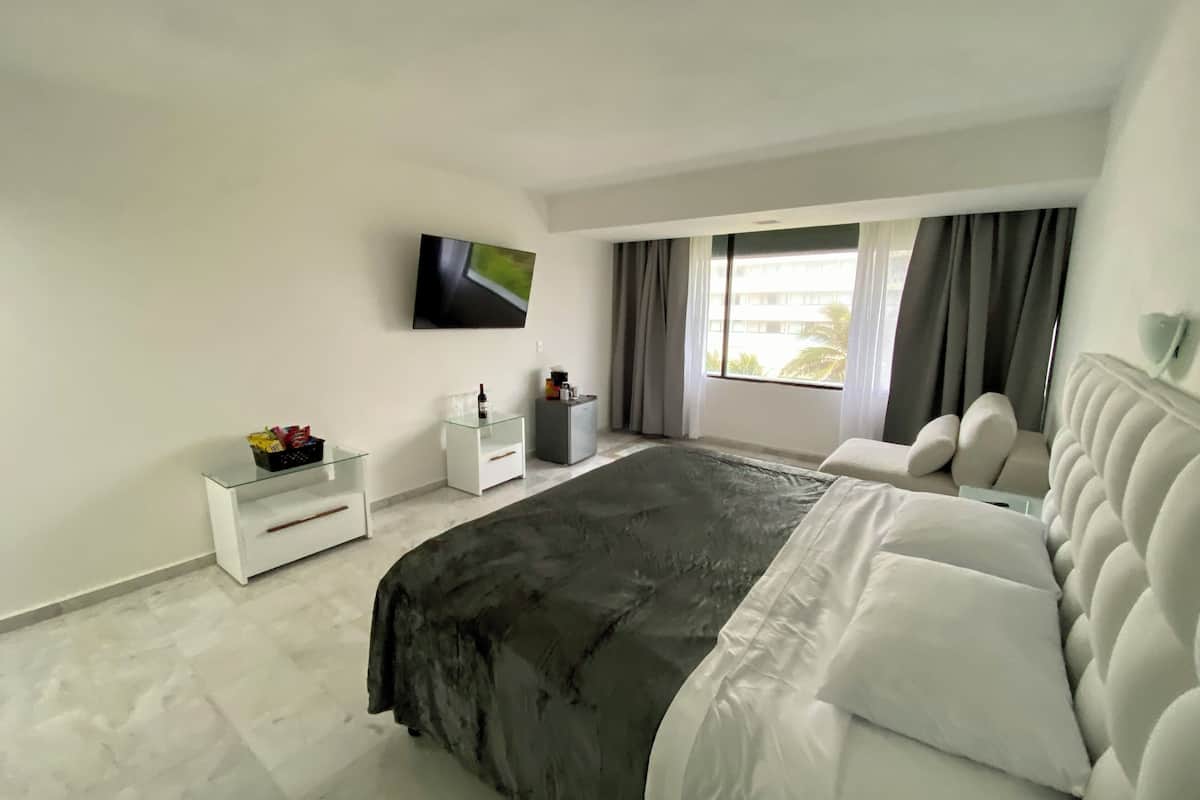 Quarto moderno com uma cama grande, uma televisão na parede, uma mesa de cabeceira com uma garrafa e uma janela com cortinas.