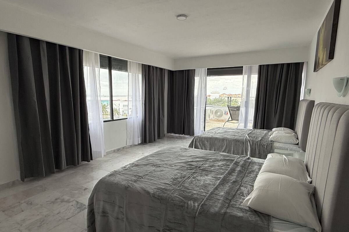 Quarto de hotel com duas camas, janelas amplas com cortinas, um ventilador de teto e vista para o exterior.