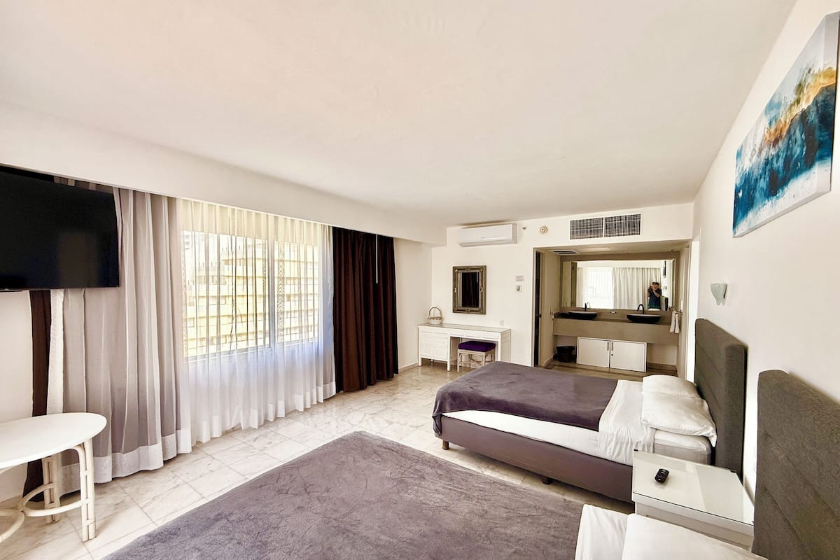 Quarto de hotel com duas camas, uma escrivaninha, televisão, banheiro com pia e espelho, e um quadro na parede.