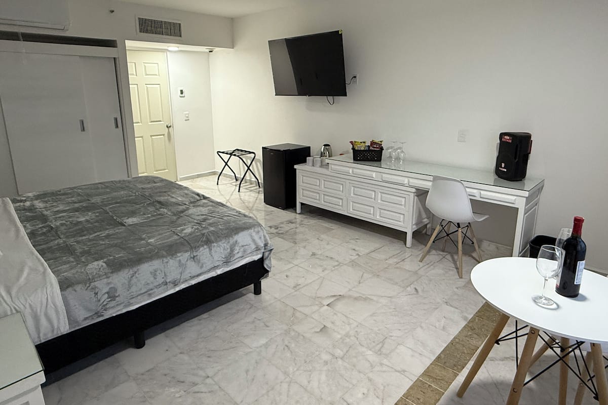 Quarto de hotel com uma cama, uma secretária com cadeira, uma mesinha com uma taça de vinho e uma televisão na parede.