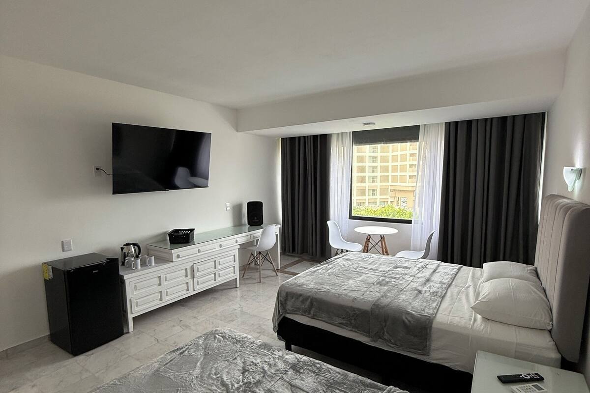 Quarto de hotel com duas camas, uma escrivaninha, televisão de tela plana, minibar e janela com cortinas.