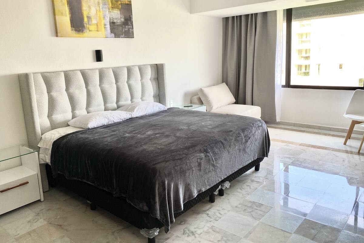 Quarto moderno com uma cama grande, mesa de cabeceira de vidro, uma cadeira e vista para edifícios pela janela.