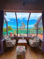 Um quarto com piso de madeira, duas cadeiras com padrão floral, uma mesa pequena com abajur e vista para o oceano por grandes janelas.
