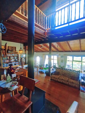 Um espaço amplo com teto alto, vigas de madeira, área de refeições com cadeiras e mesa, sala de estar com sofá e uma estante repleta de livros.