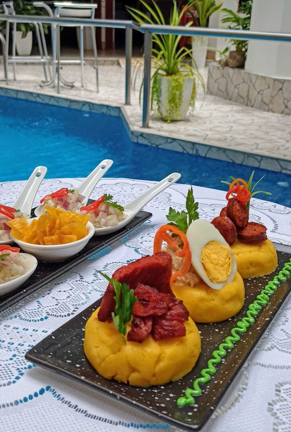 Disposição de refeições junto à piscina, com diversos pratos, incluindo um item empanado amarelo coberto com carne e ovo, e recipientes com batatas fritas e salsa.