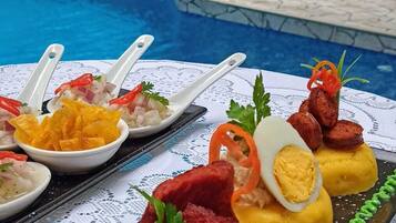 Disposição de refeições junto à piscina, com diversos pratos, incluindo um item empanado amarelo coberto com carne e ovo, e recipientes com batatas fritas e salsa.