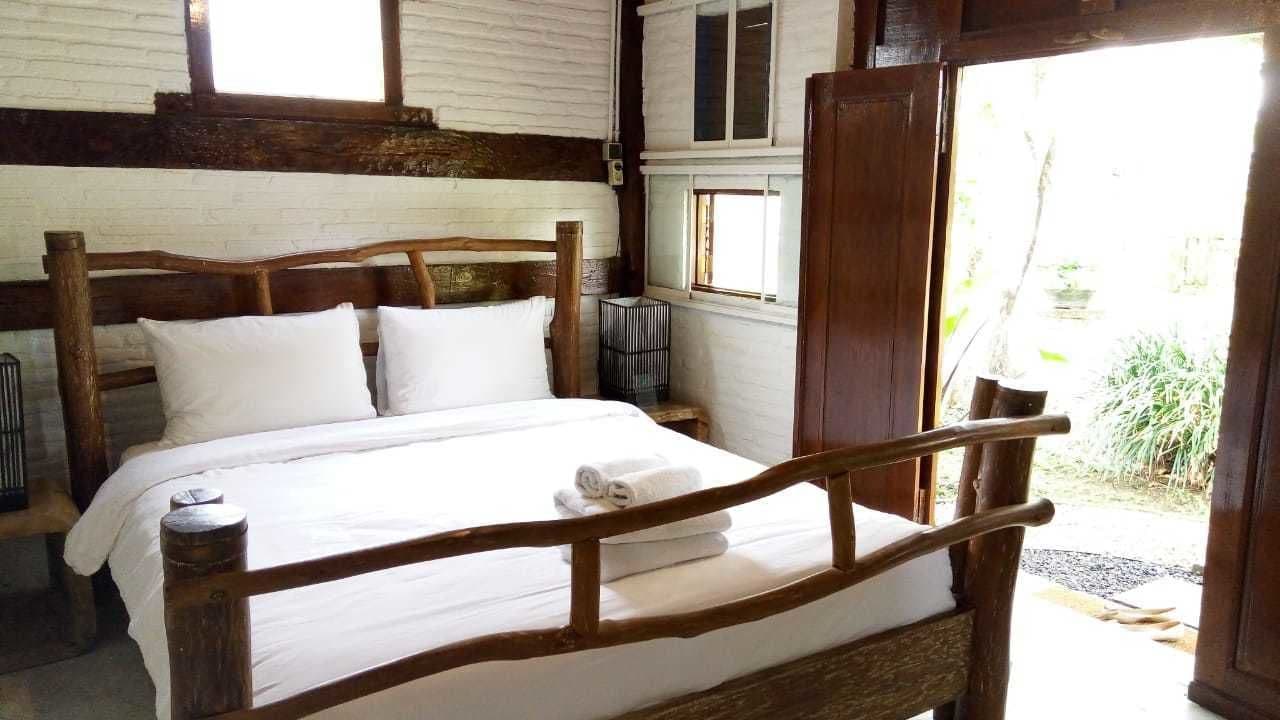 Uma cama de madeira com roupa de cama branca e uma toalha enrolada, uma pequena mesa de cabeceira e uma janela com vista para vegetação.