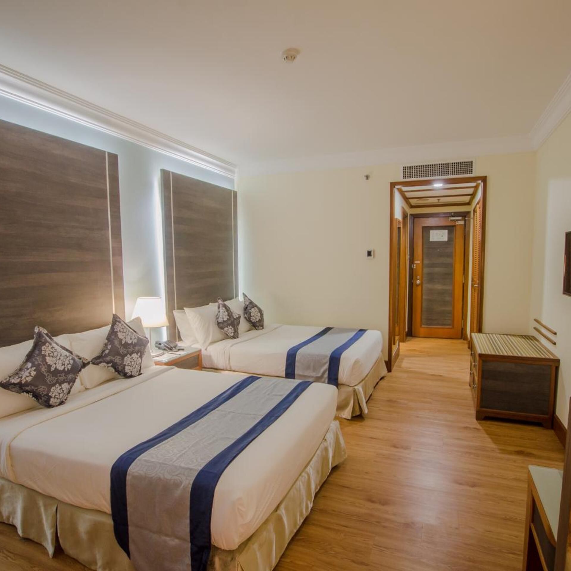 Quarto de hotel com duas camas, piso de madeira, uma escrivaninha e uma porta que dá para outro espaço.