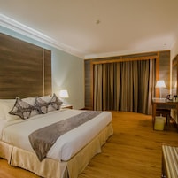 Quarto de hotel com uma cama grande, uma escrivaninha com cadeira, um abajur e uma cortina.