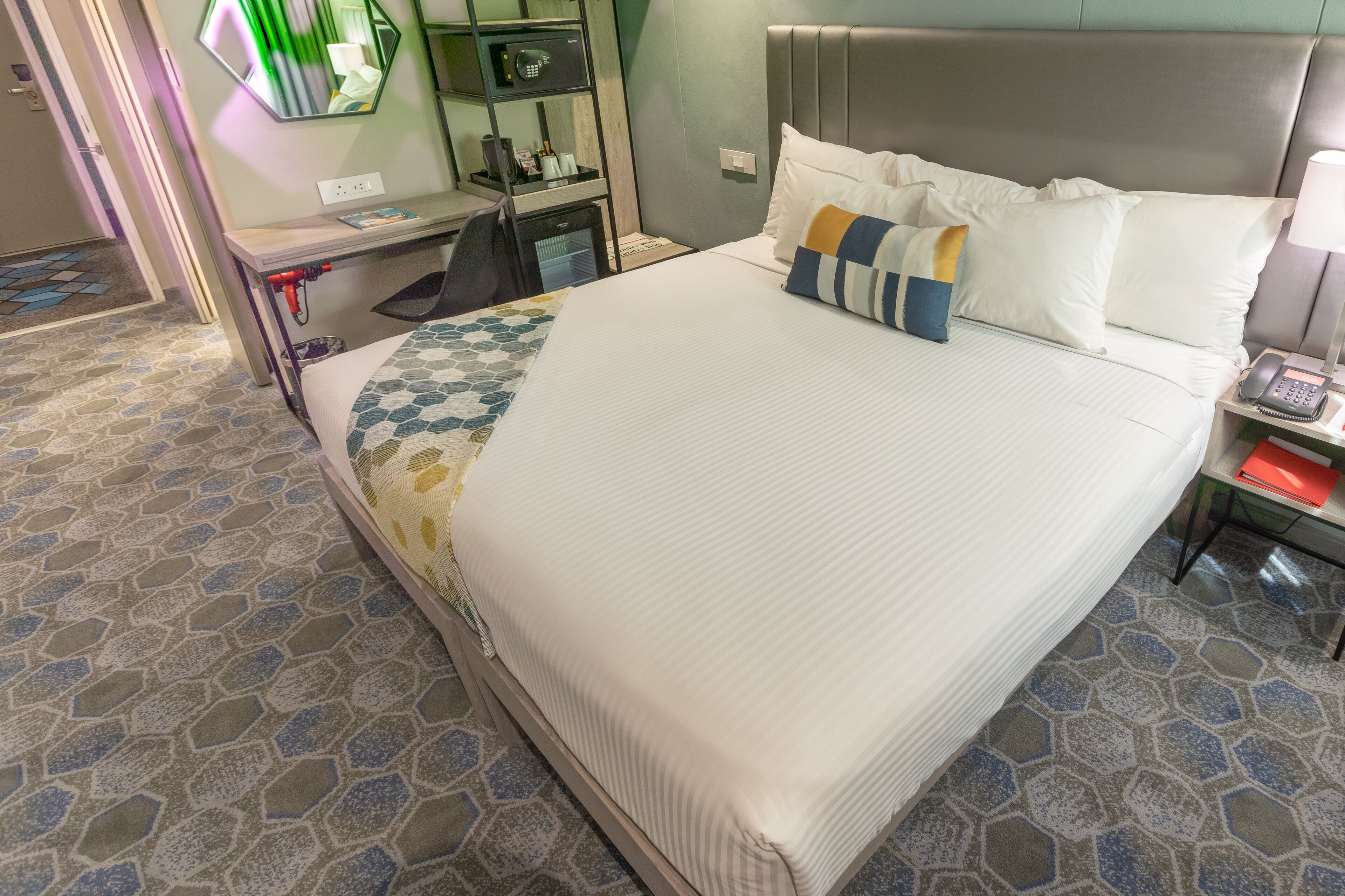 Cama bem arrumada com travesseiro listrado, mesa de cabeceira com telefone e escrivaninha com cadeira e abajur.