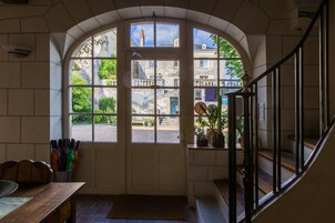 Uma janela arqueada com vários painéis, um banco de madeira, um recipiente de metal para guarda-chuvas e uma escadaria que leva a um andar superior.