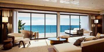Quarto de hotel moderno com uma varanda ampla, um sofá, uma mesa de centro redonda e vista para o oceano.