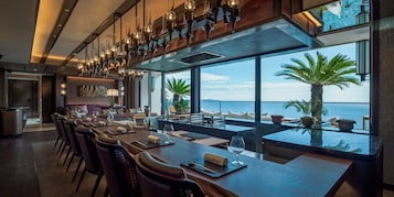 Um restaurante com uma mesa de jantar comprida, cadeiras e vista para o mar.