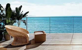 Uma poltrona e um banquinho de vime em um deck revestido com azulejos, com vista para o oceano.
