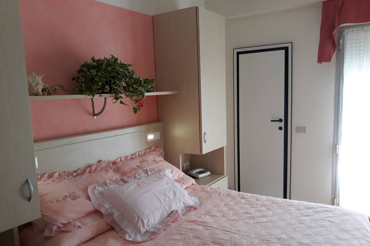 Quarto com colcha rosa, travesseiro branco, prateleira com planta e porta com puxador.