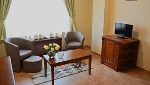 Uma sala de estar com dois poltronas, uma mesa de centro com um vaso de flores, uma televisão sobre um aparador de madeira e uma janela com cortinas.