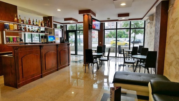 Uma área de bar com balcão, prateleiras cheias de garrafas e uma televisão. Mesas e cadeiras dispostas para acomodar clientes.