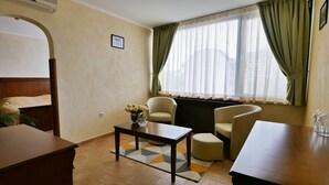 Quarto de hotel com uma cama, uma mesinha com um vaso de flores, duas poltronas, uma janela com cortinas e um radiador.