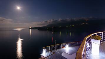 Vista noturna de uma varanda com vista para águas calmas, uma linha de costa distante e uma lua brilhante.