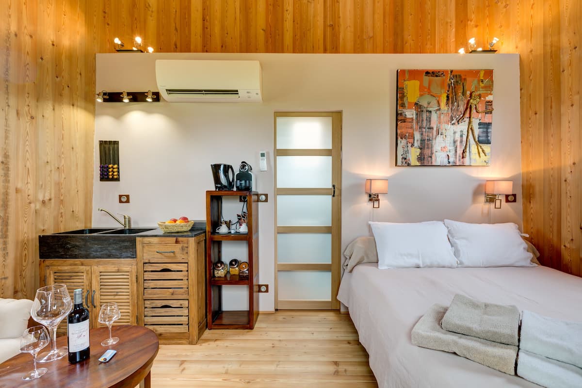 Quarto de hotel compacto com decoração em madeira, cama com roupa de cama branca, pequena cozinha e um quadro na parede.