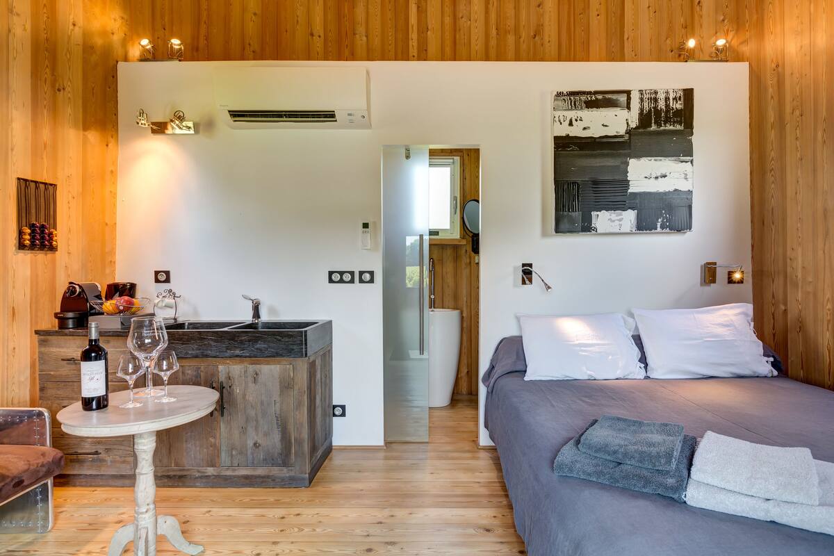Quarto moderno com teto de madeira, cama com roupa de cama branca e cinza, pequena cozinha com geladeira e mesa de jantar com copos de vinho e uma garrafa.