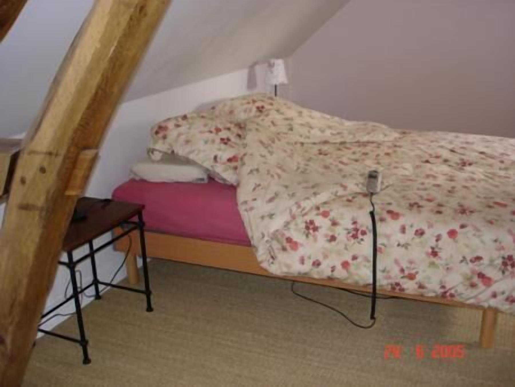 Quarto pequeno com uma cama de solteiro, colcha floral, mesa de cabeceira e abajur.