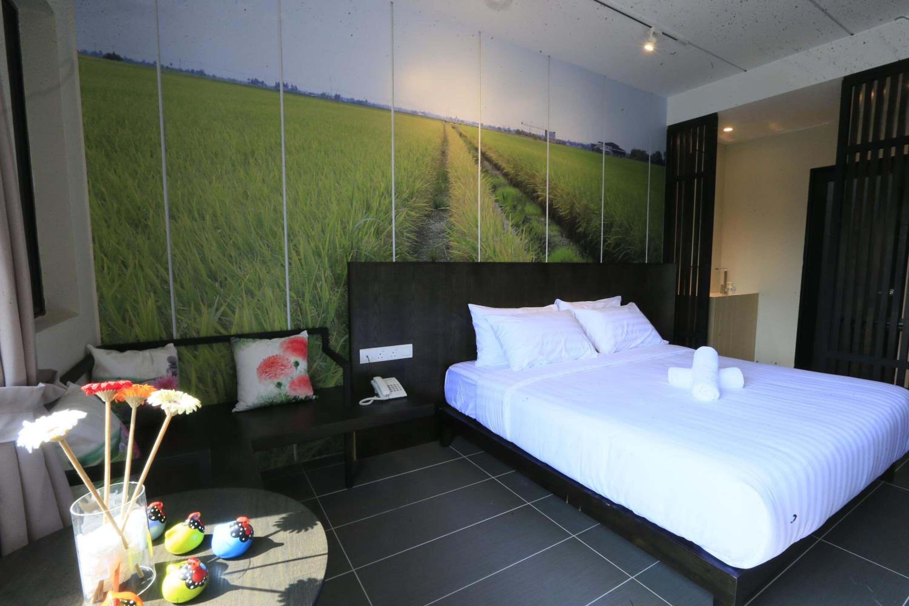 Quarto de hotel com uma cama grande, uma mesa de cabeceira, uma pequena mesa com objetos decorativos e um mural na parede retratando um campo.