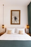 Cama bem arrumada com lençóis brancos e duas almofadas, um quadro com uma palmeira na parede e duas luminárias fixadas nas paredes, de cada lado.