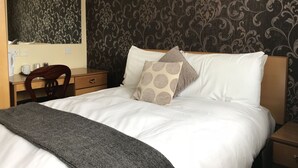 Cama bem arrumada com lençóis brancos, manta cinza e almofadas estampadas. Há uma mesinha de cabeceira de madeira com um abajur, uma cadeira e uma pequena mesa com uma xícara e uma garrafa. A parede apresenta um papel de parede escuro e ornamental.