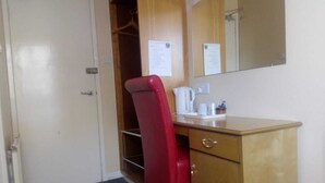 Quarto de hotel com uma escrivaninha de madeira, uma cadeira vermelha, uma cama branca, um espelho e uma porta.