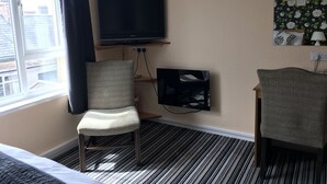 Quarto de hotel com cama, televisão sobre suporte, uma cadeira, mesa pequena e janela com cortinas.