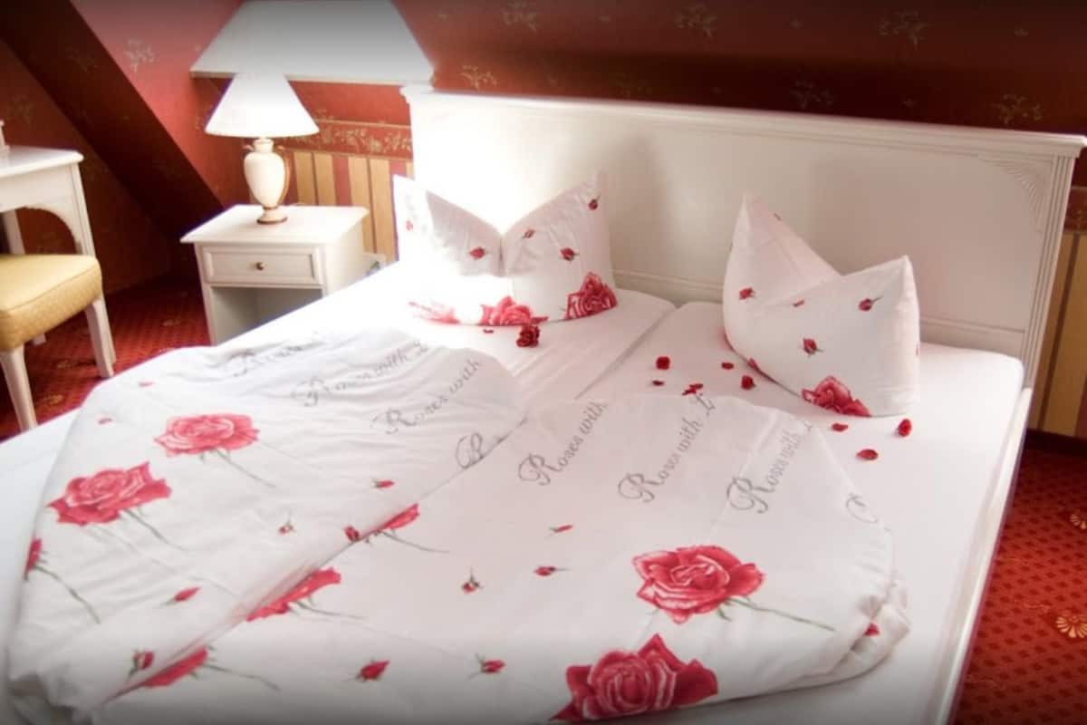 Cama bem arrumada com roupa de cama e almofadas florais, mesa de cabeceira com abajur e uma cadeira.