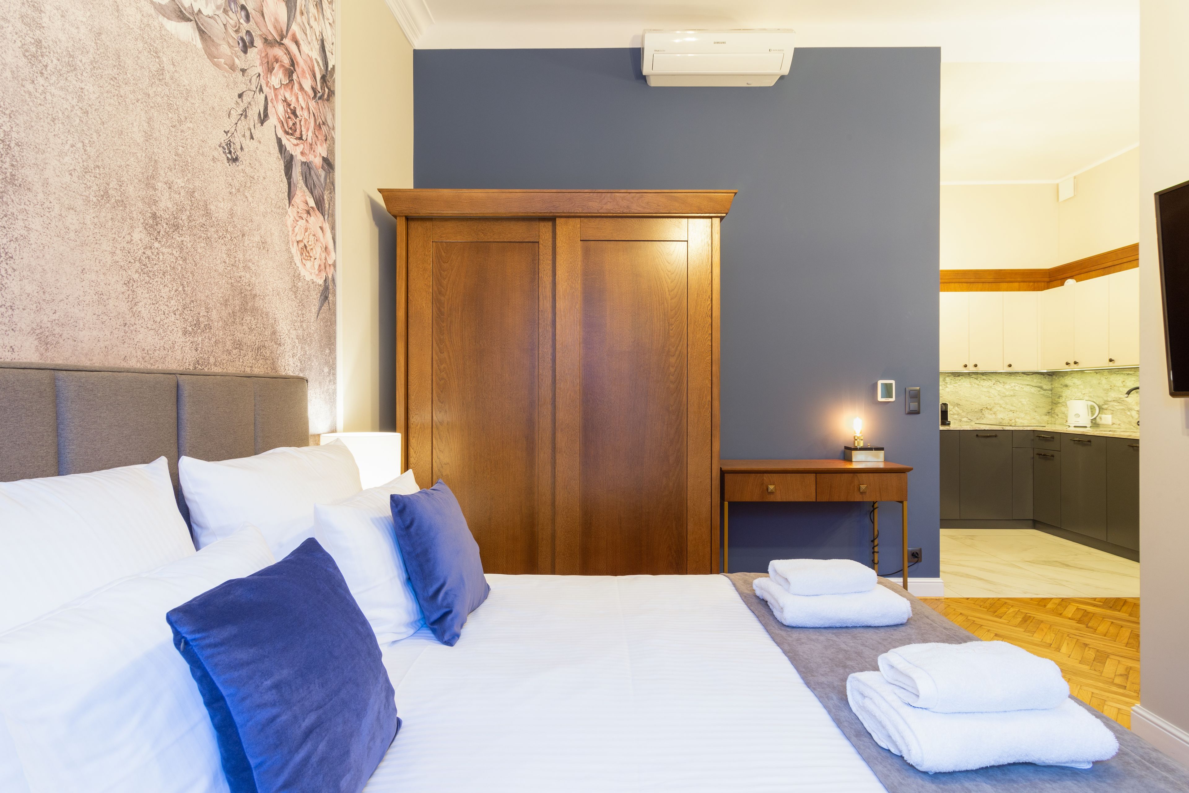 Quarto de hotel com uma cama grande, um guarda-roupa de madeira, uma pequena escrivaninha com luminária e vista para a cozinha.
