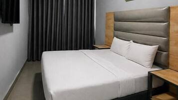 Quarto de hotel padrão com cama, cabeceira, mesa de cabeceira, televisão e cortina.