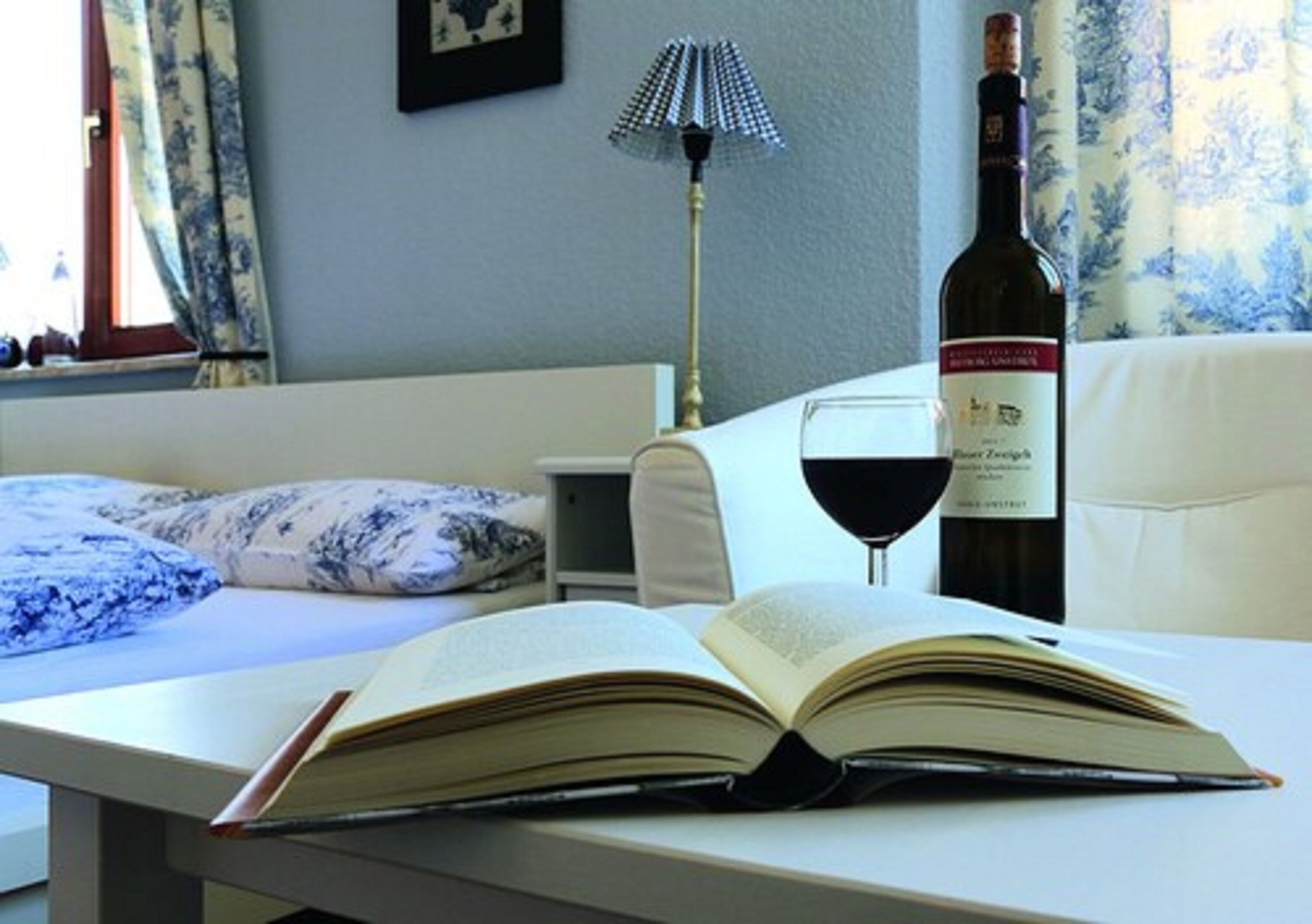 Um quarto com uma cama, um livro aberto, uma garrafa de vinho e um copo de vinho sobre uma mesa de cabeceira.
