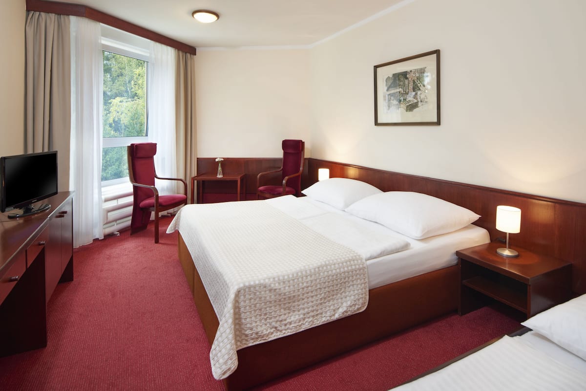Quarto de hotel com uma cama grande, uma escrivaninha com televisão, duas cadeiras vermelhas, uma mesa de cabeceira com abajur e um quadro pendurado na parede.