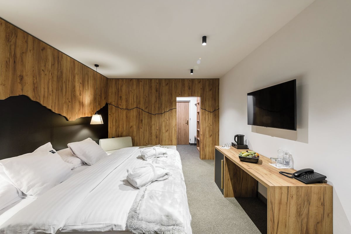 Quarto de hotel moderno com cabeceira de madeira, uma cama grande com roupa de cama branca, uma escrivaninha com cafeteira e uma TV de tela plana fixada na parede.