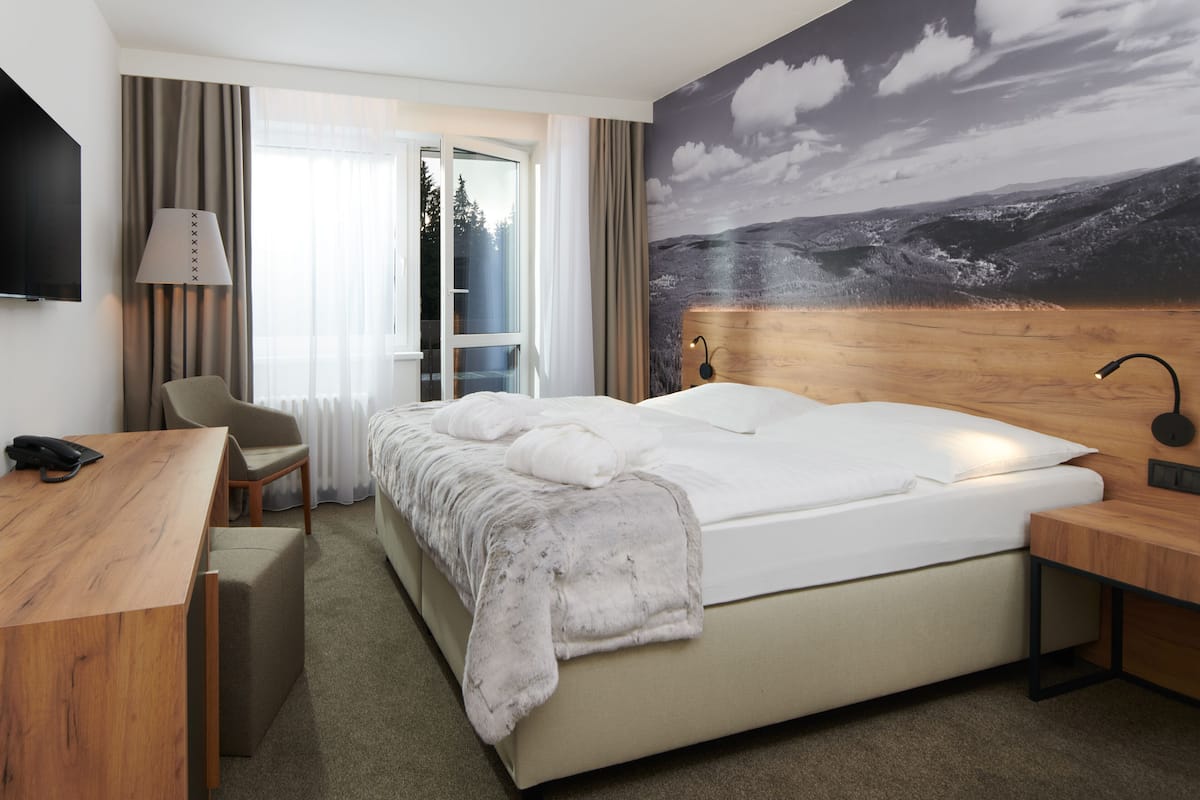 Quarto de hotel com cama, escrivaninha, cadeira, luminária e uma grande pintura de paisagem na parede.