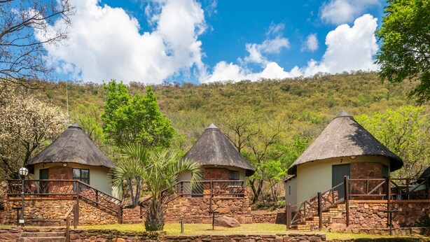 Três cabanas com telhados de palha e paredes de pedra, uma escadaria de pedra e um primeiro plano com grama.