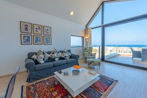 Uma sala de estar moderna com um sofá cinzento, uma mesa de centro e uma janela ampla com vista para o mar.