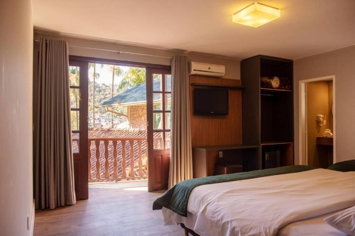 Quarto de hotel com cama, televisão, escrivaninha e varanda com vista para palmeiras.