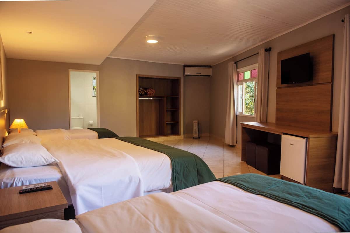 Quarto de hotel com três camas, televisão, guarda-roupa e secretária com abajur.
