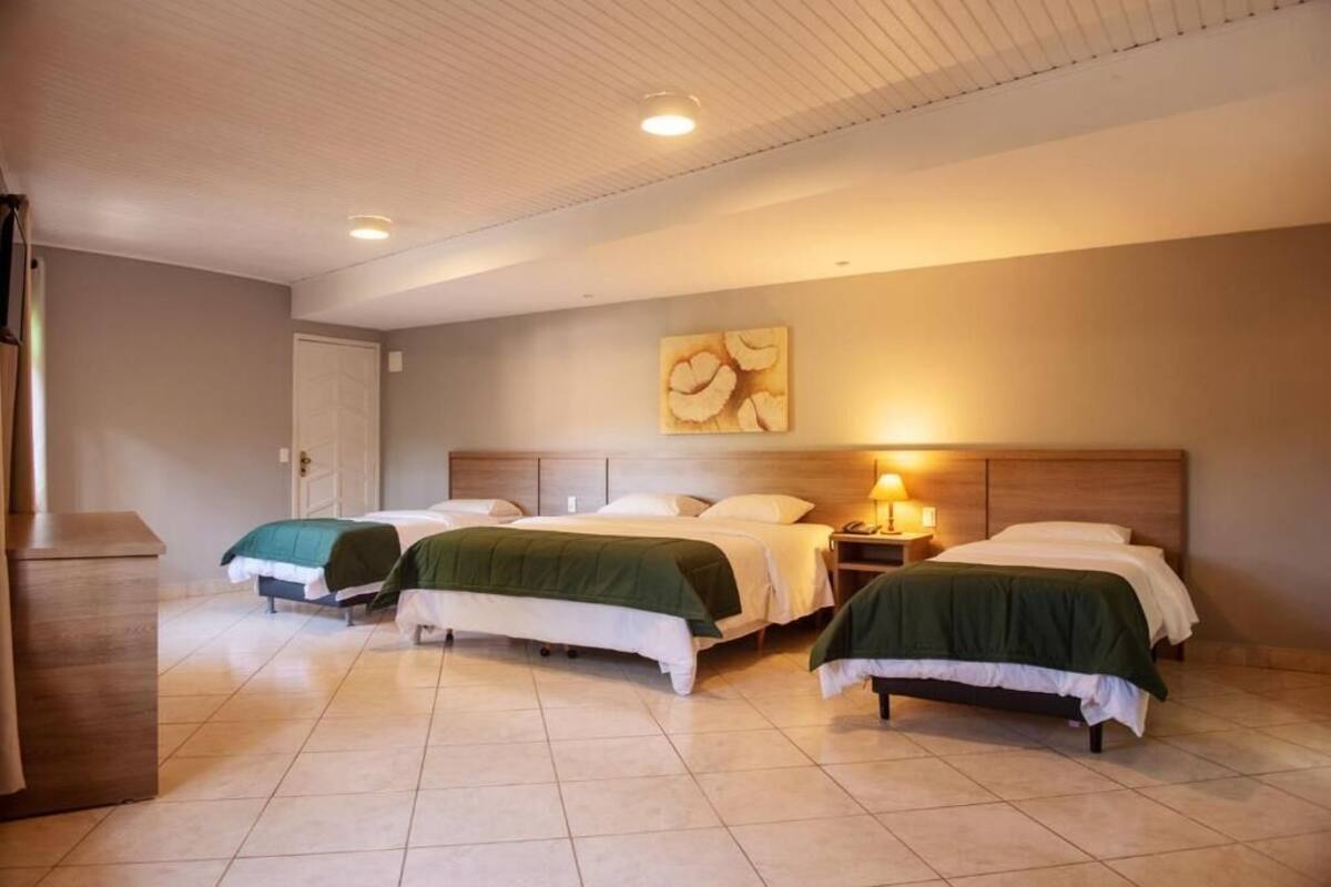 Quarto de hotel com três camas, cabeceira de madeira, mesa de cabeceira com abajur e uma obra de arte na parede.