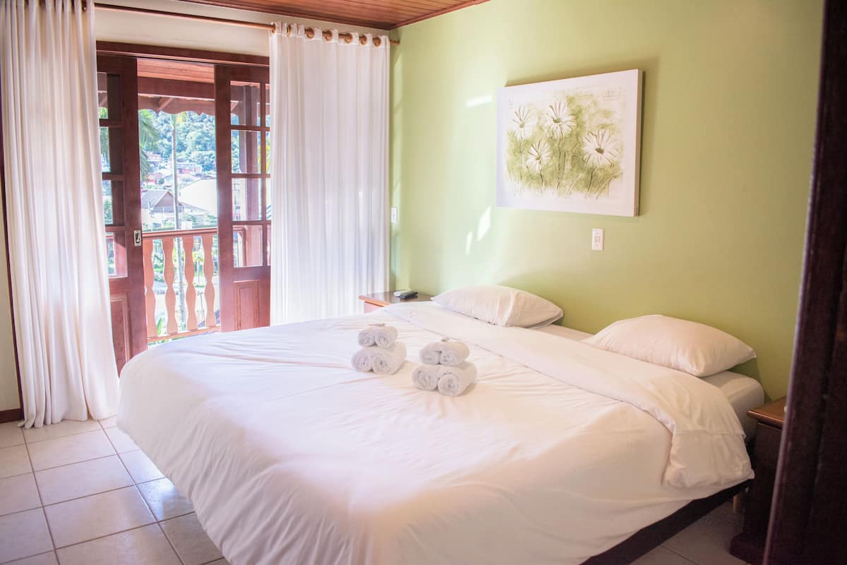 Quarto de hotel com cama, roupa de cama branca, um quadro na parede e uma varanda com cortinas.