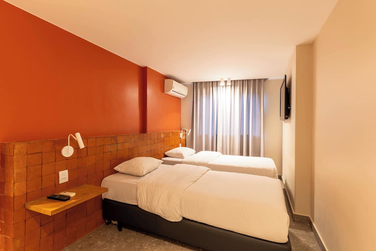 Quarto de hotel com duas camas, cabeceira de madeira, mesa de cabeceira com telefone e televisão fixada na parede.
