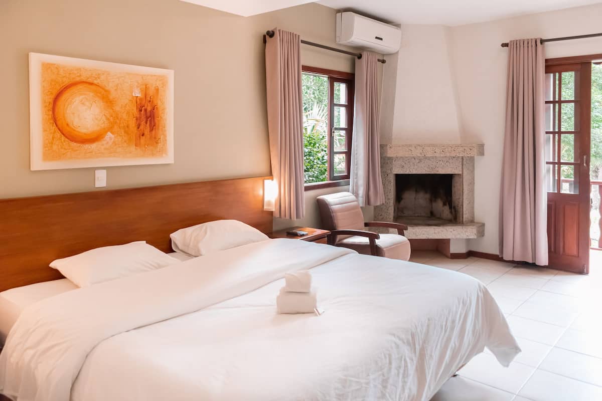 Quarto de hotel com uma cama grande, lareira, janela com cortinas e uma obra de arte pendurada na parede.